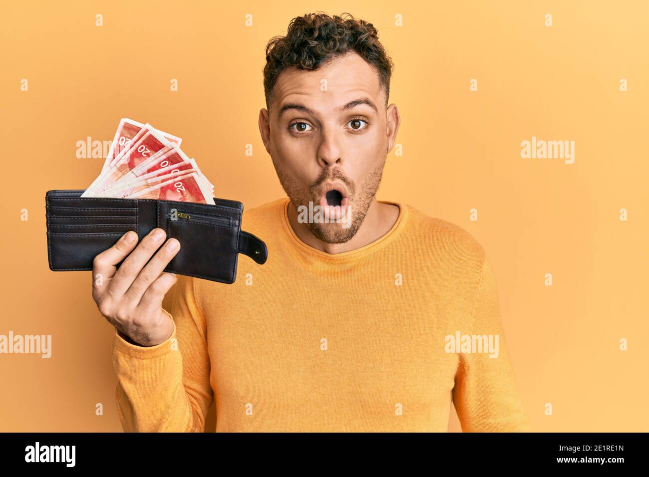 Young hispanic man holding wallet with israel shekels scared and amazed ...