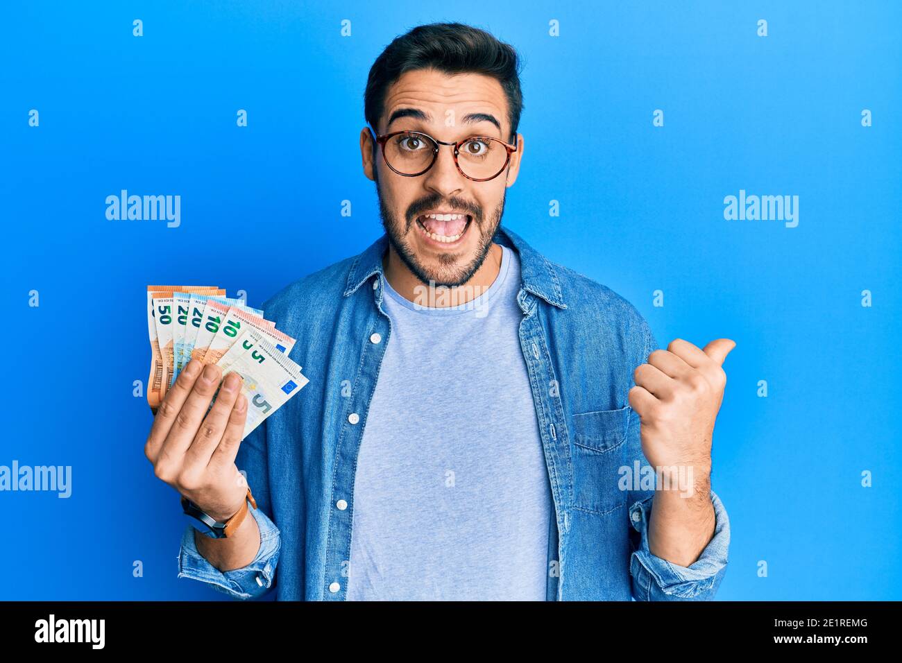 Young hispanic man holding euro banknotes pointing thumb up to the side ...