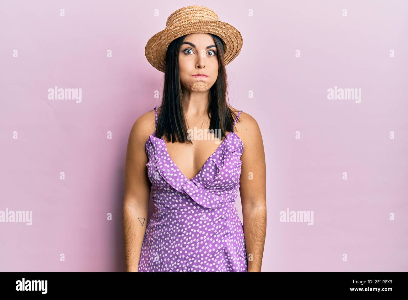 Young brunette woman wearing summer dress and hat puffing cheeks with ...