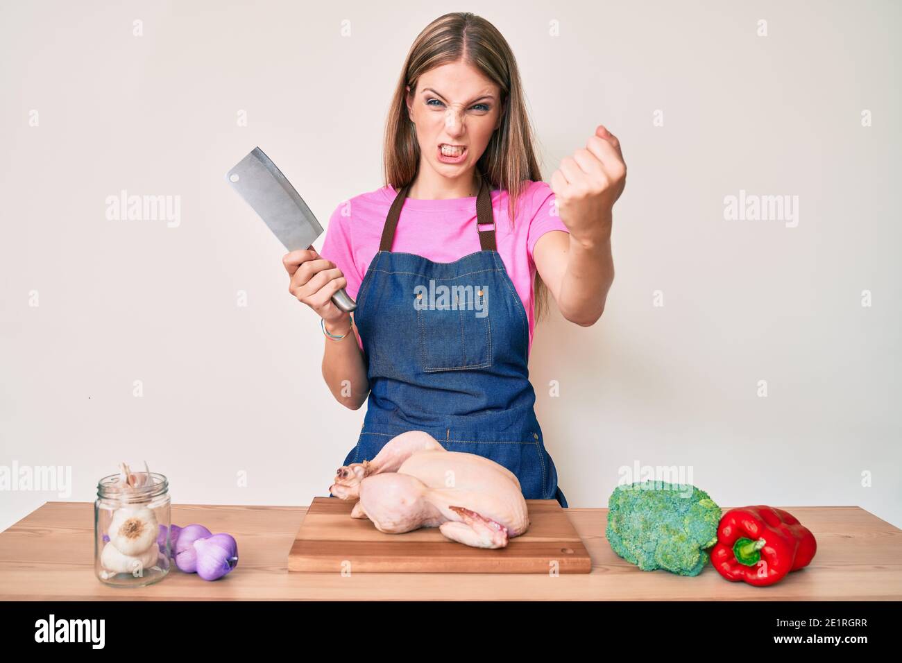 Young blonde girl wearing cook apron cooking chicken annoyed and ...
