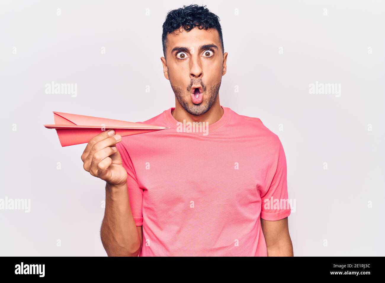 Young hispanic man holding paper airplane scared and amazed with open ...