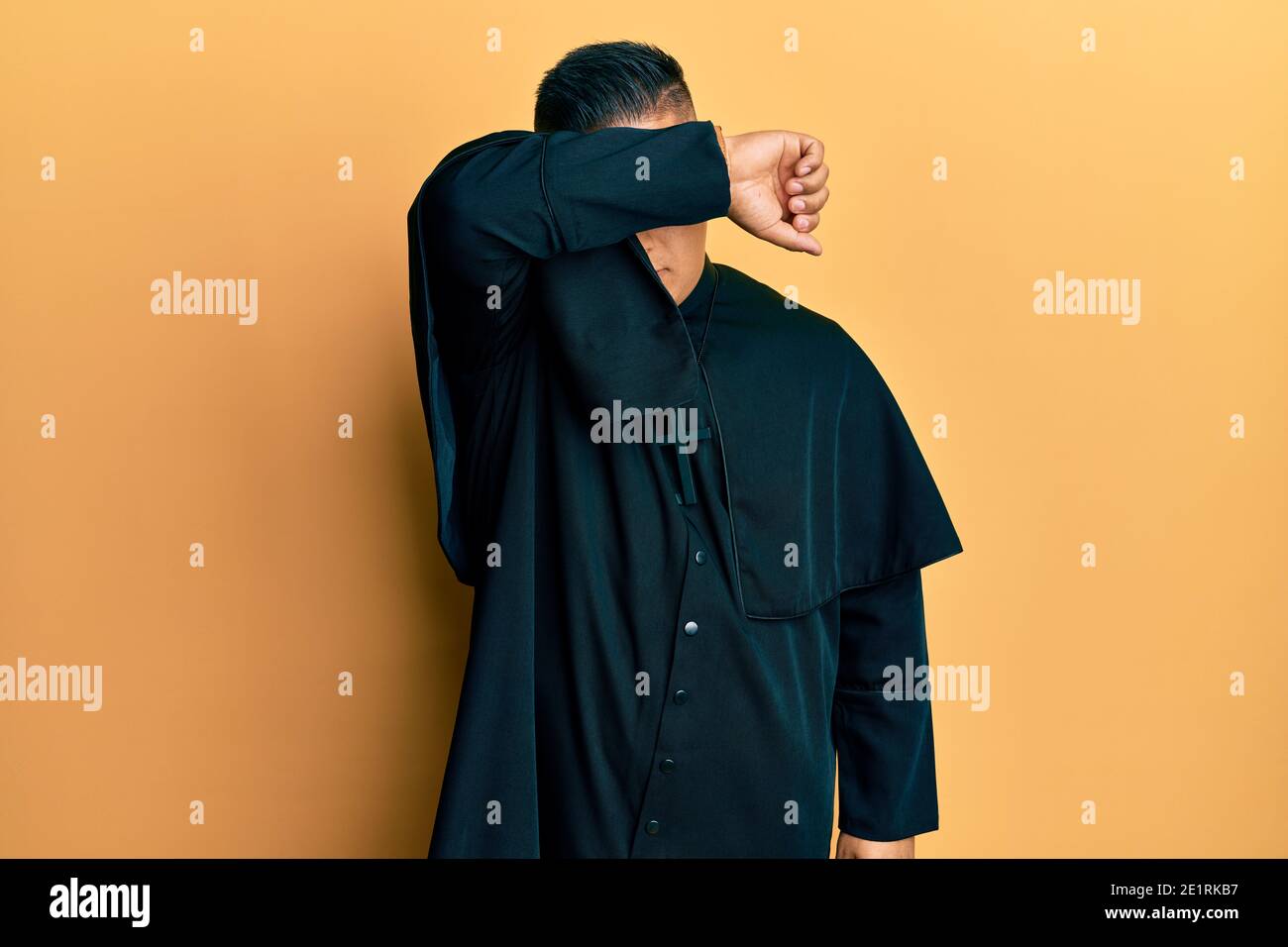 Young latin priest man standing over yellow background covering eyes ...