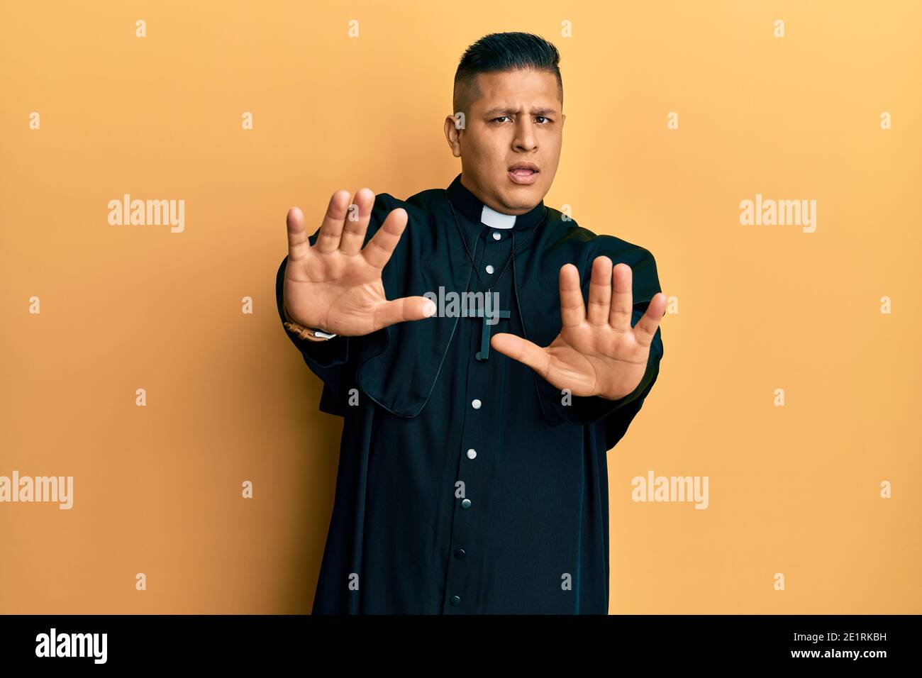 Young latin priest man standing over yellow background doing stop ...