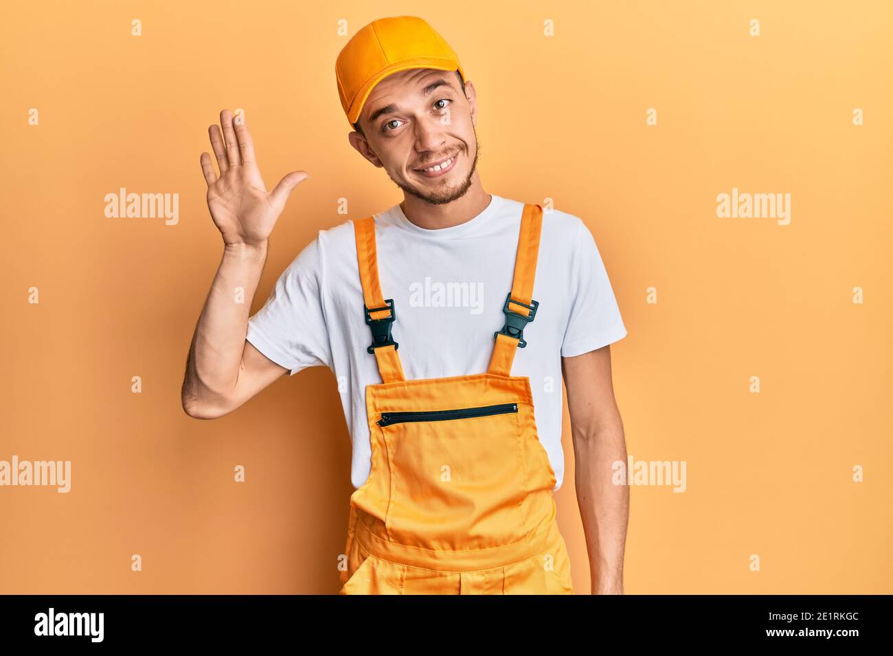 Hispanic young man wearing handyman uniform waiving saying hello happy ...