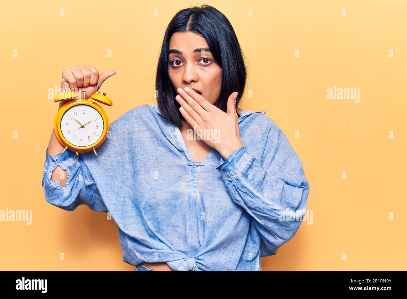 Young beautiful latin woman holding alarm clock covering mouth with ...
