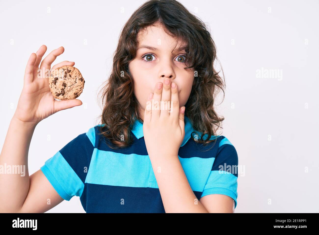 Cute hispanic child with long hair holding cookie covering mouth with ...