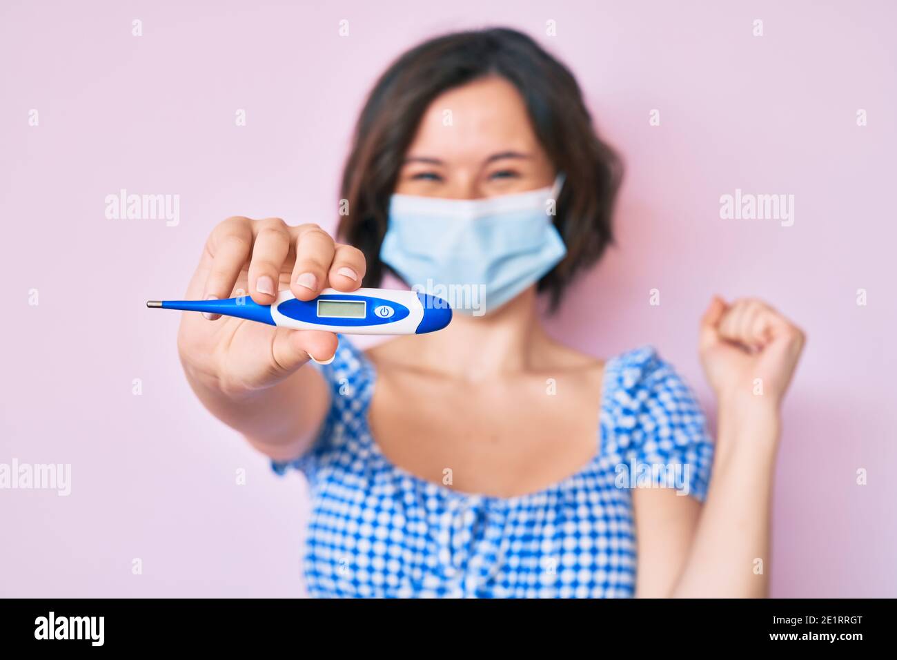Young beautiful woman wearing medical mask holding holding thermometer ...