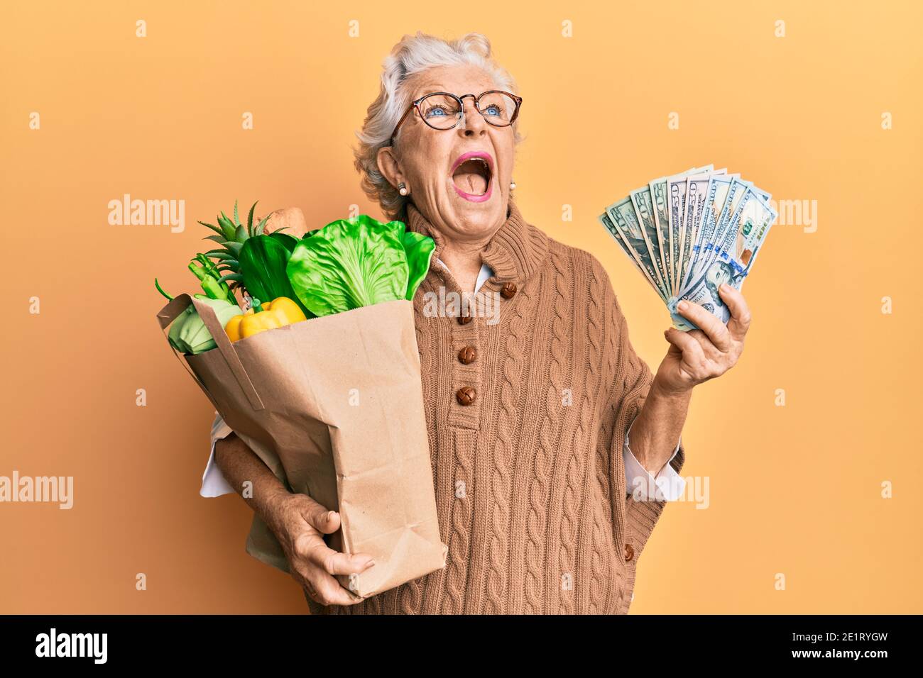Senior grey-haired woman holding groceries and united states dollars ...