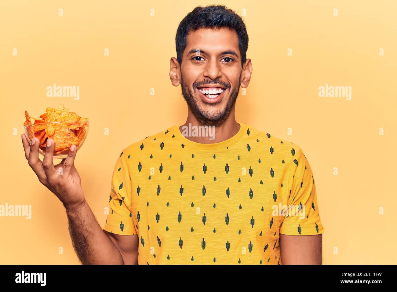 Young latin man holding nachos potato chips looking positive and happy ...