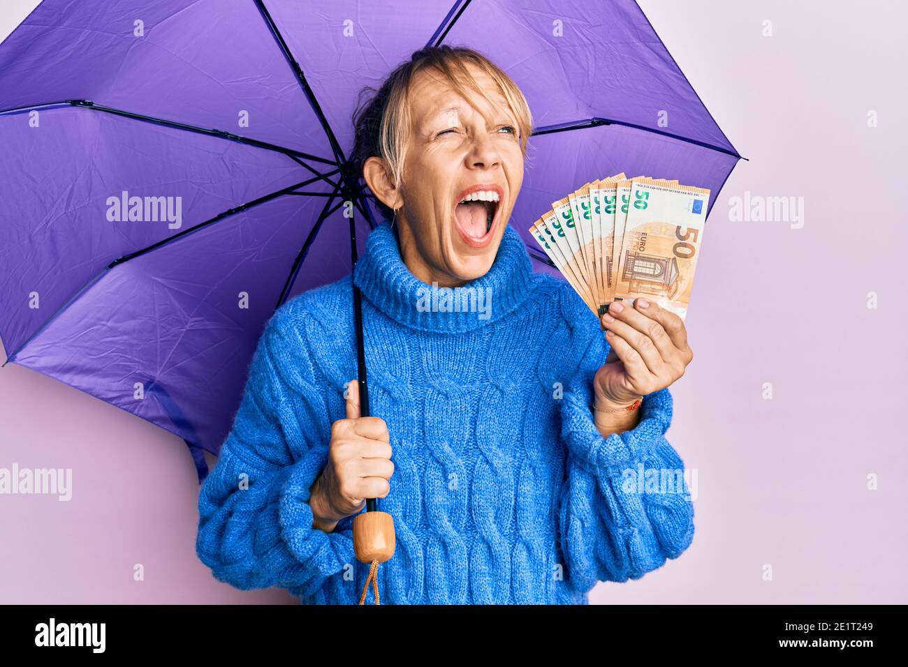 Middle age blonde woman holding umbrella and 50 euros banknotes angry ...