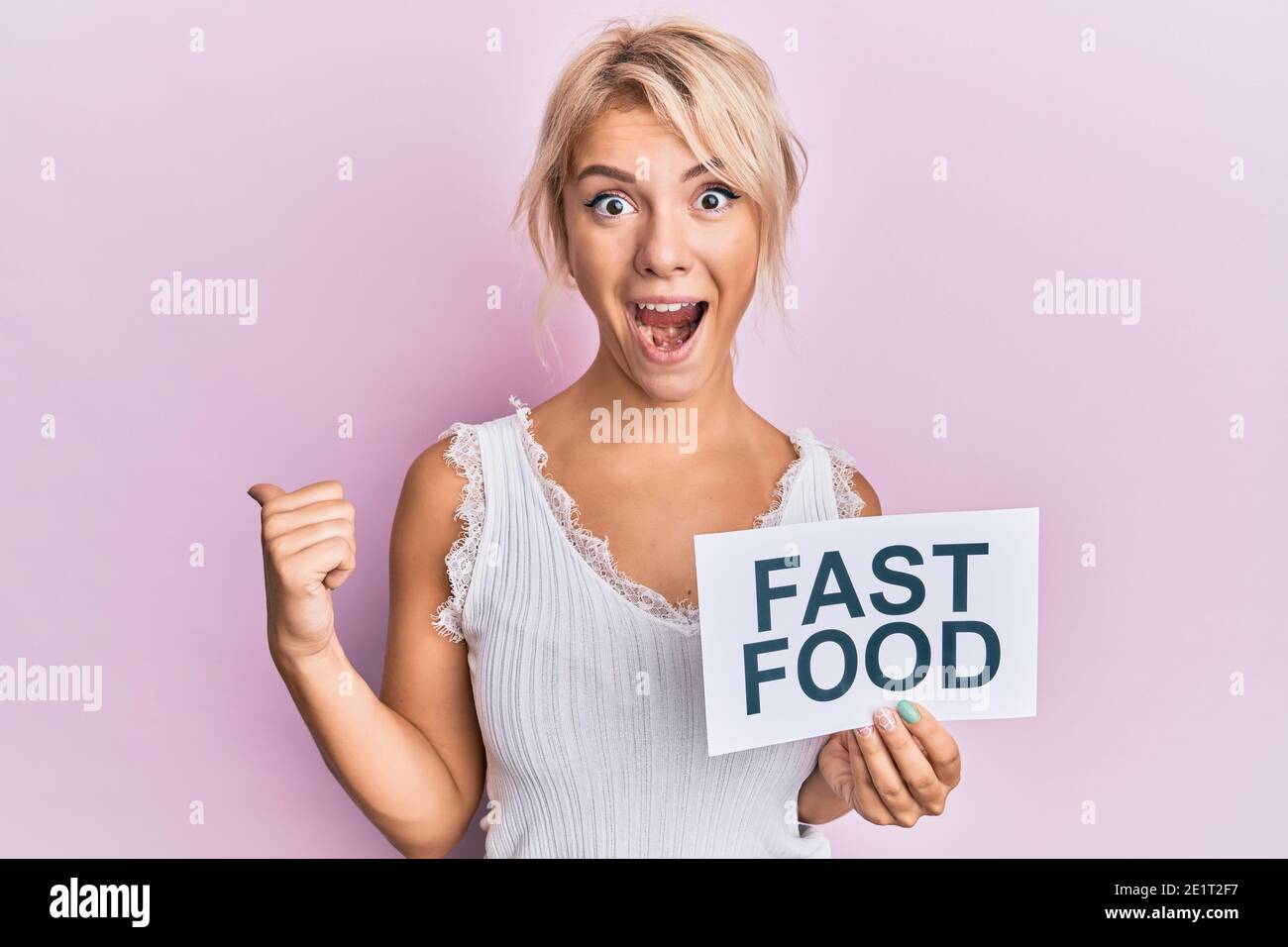 Young blonde girl eating fast food pizza and burger pointing thumb up ...