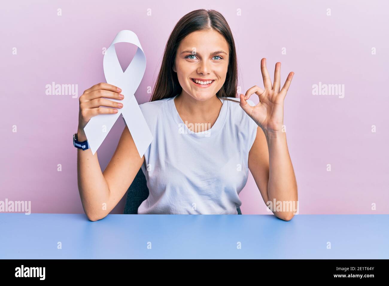 Young caucasian woman holding white ribbon doing ok sign with fingers ...
