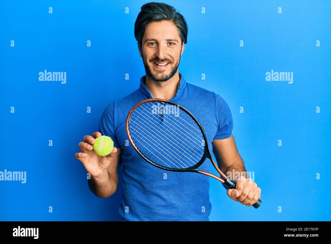 Young hispanic man playing tennis holding racket and ball smiling with ...