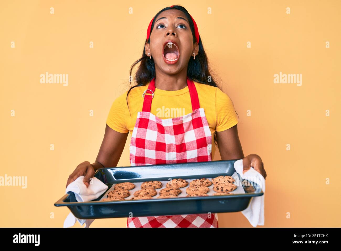 Young indian girl wearing baker uniform holding homemade cookies angry ...