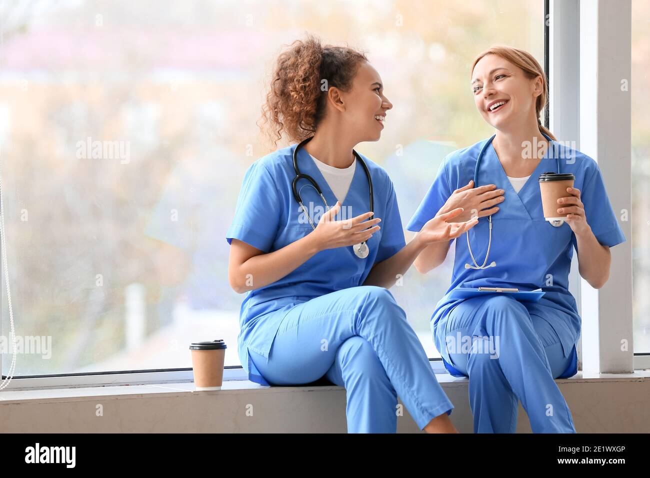 Female doctors drinking coffee while sitting on window sill in clinic ...