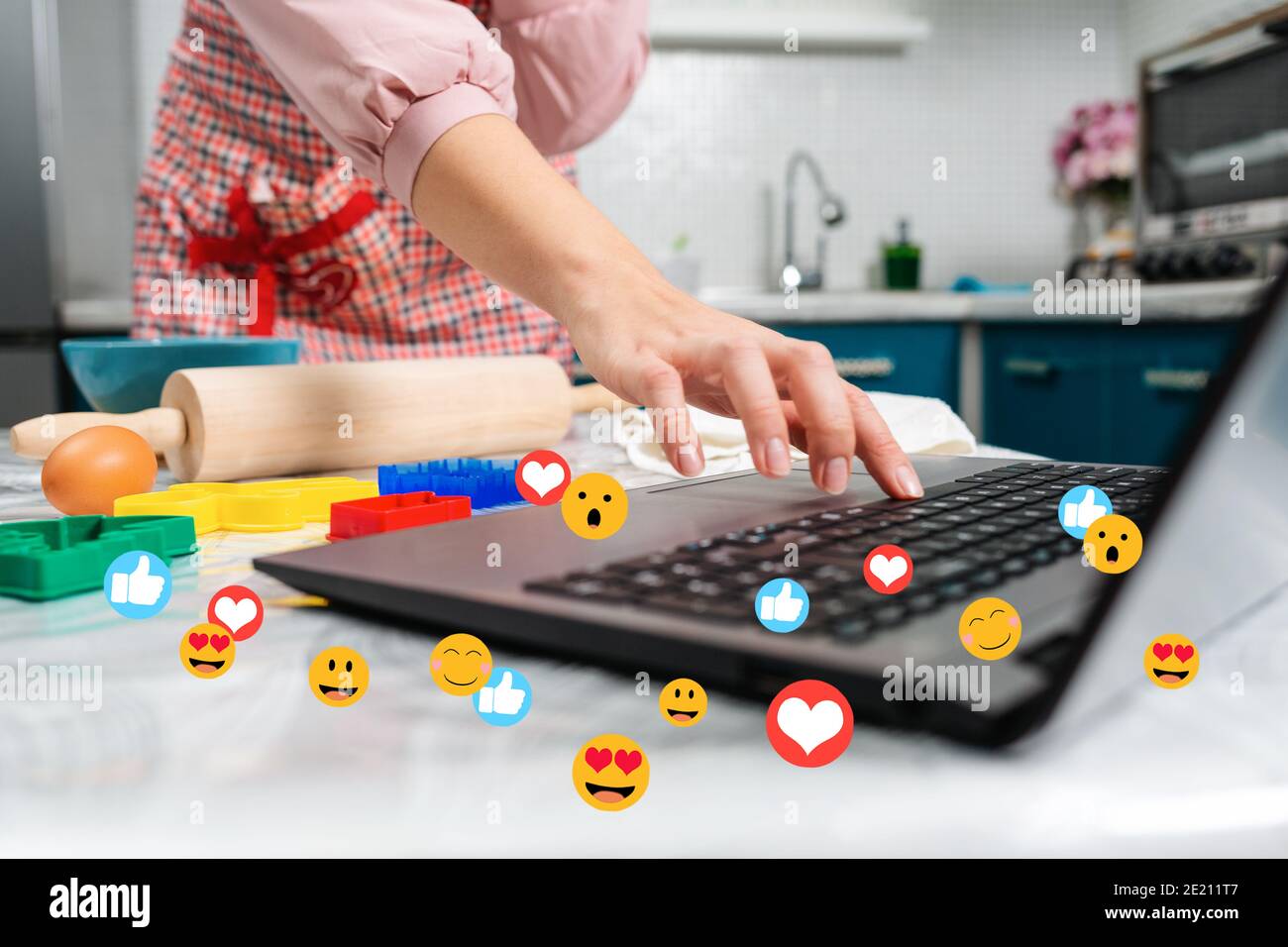 A woman in an apron uses a laptop on the kitchen table. Emoji icons fly ...