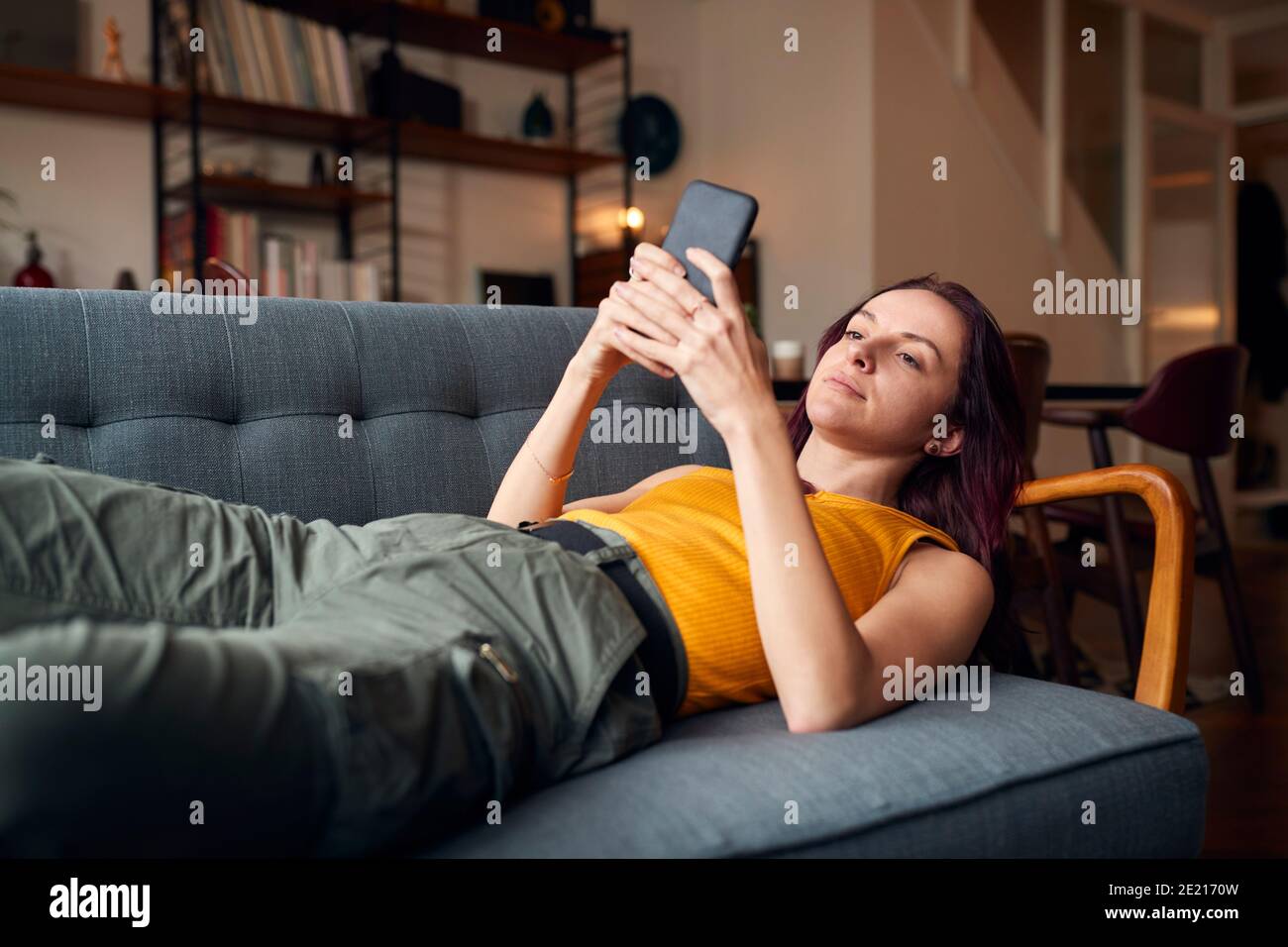 Young Woman Lying On Sofa At Home Looking At Mobile Phone Messages And ...