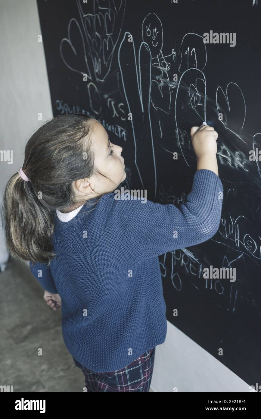 Vertical shot of a cute Caucasian little girl drawing on the blackboard ...