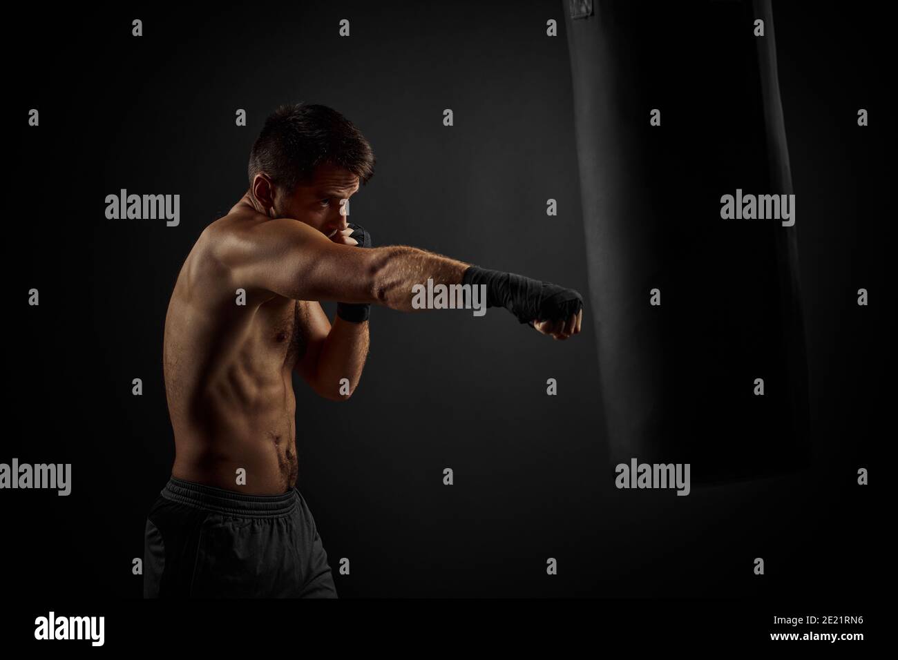 Aggressive shirtless male boxer punching in boxing bag on black ...
