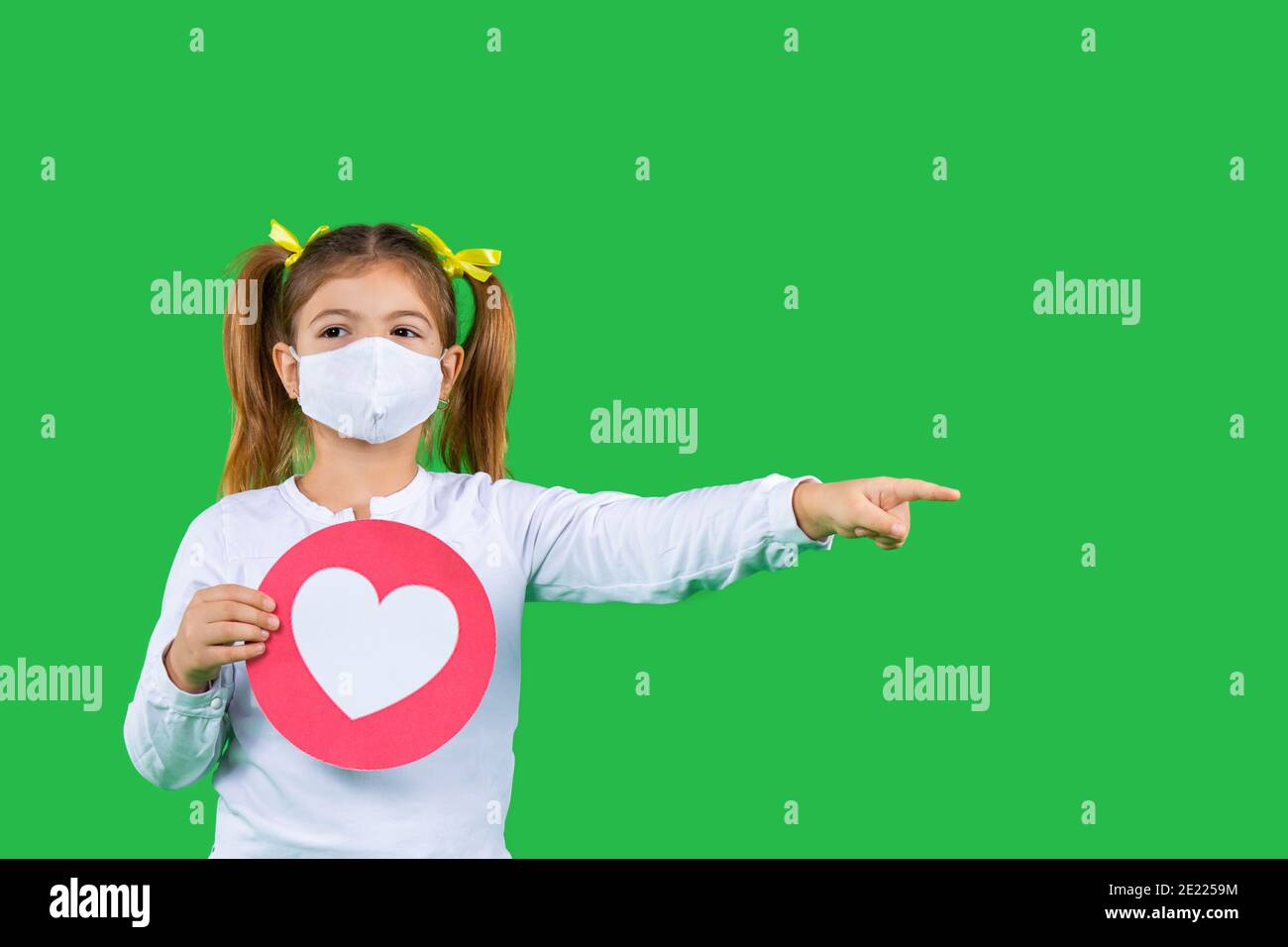 A preschool girl in a protective mask holds a sign with a red heart ...