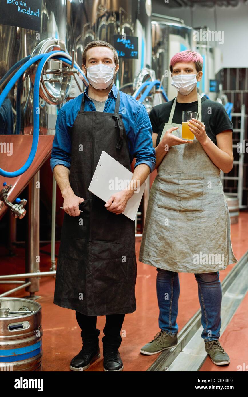 Two young brewers in workwear and protective masks standing in front of ...
