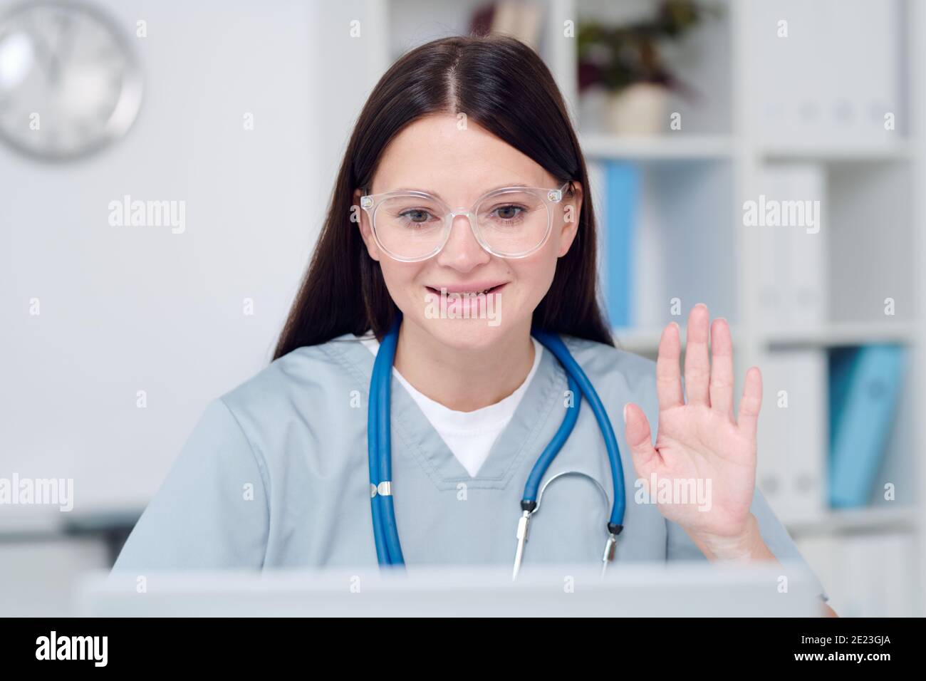 Happy young brunette doctor in uniform and eyeglasses greeting online ...