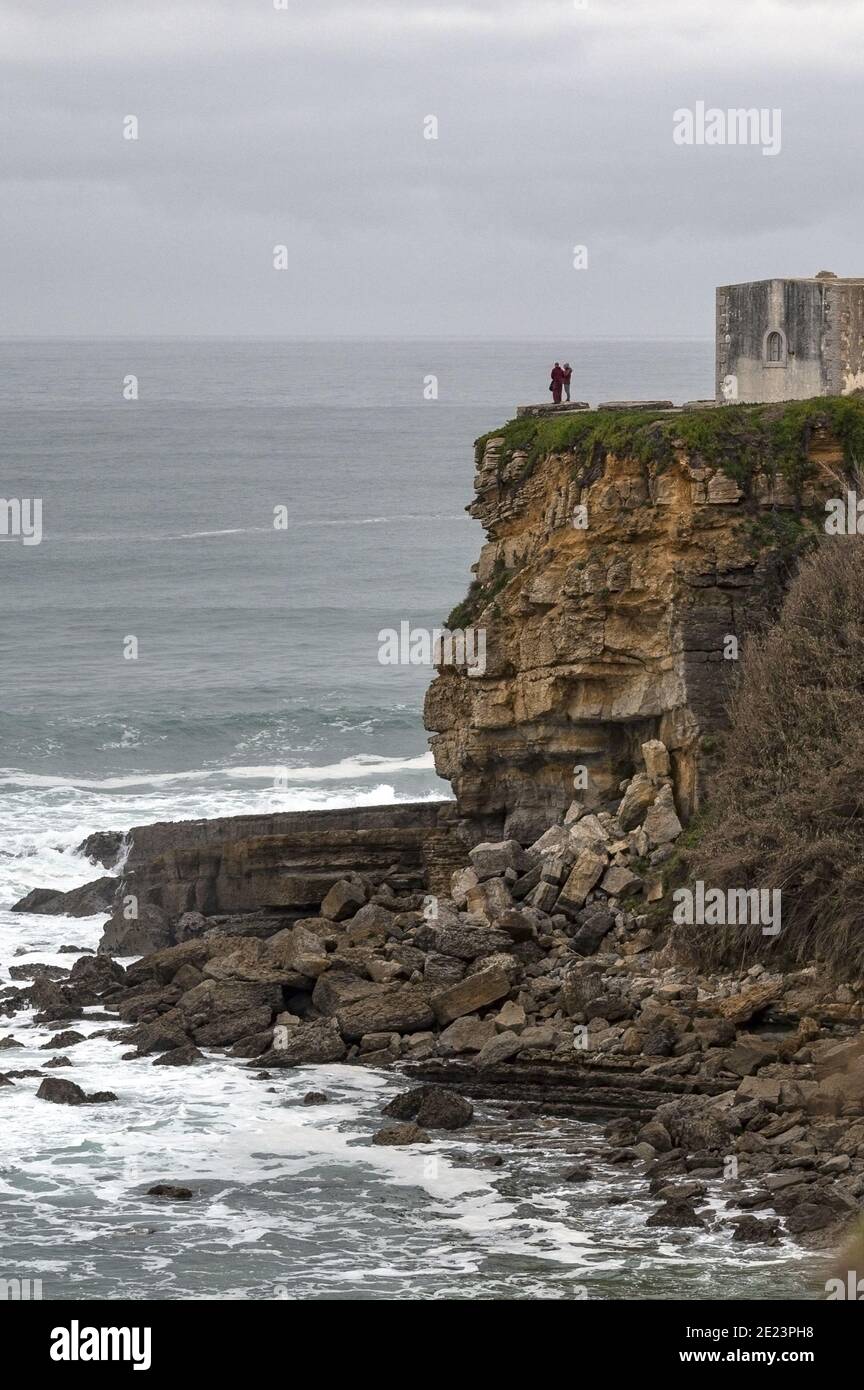 Vertical shot of a huge cliff on the beach with two people standing on ...