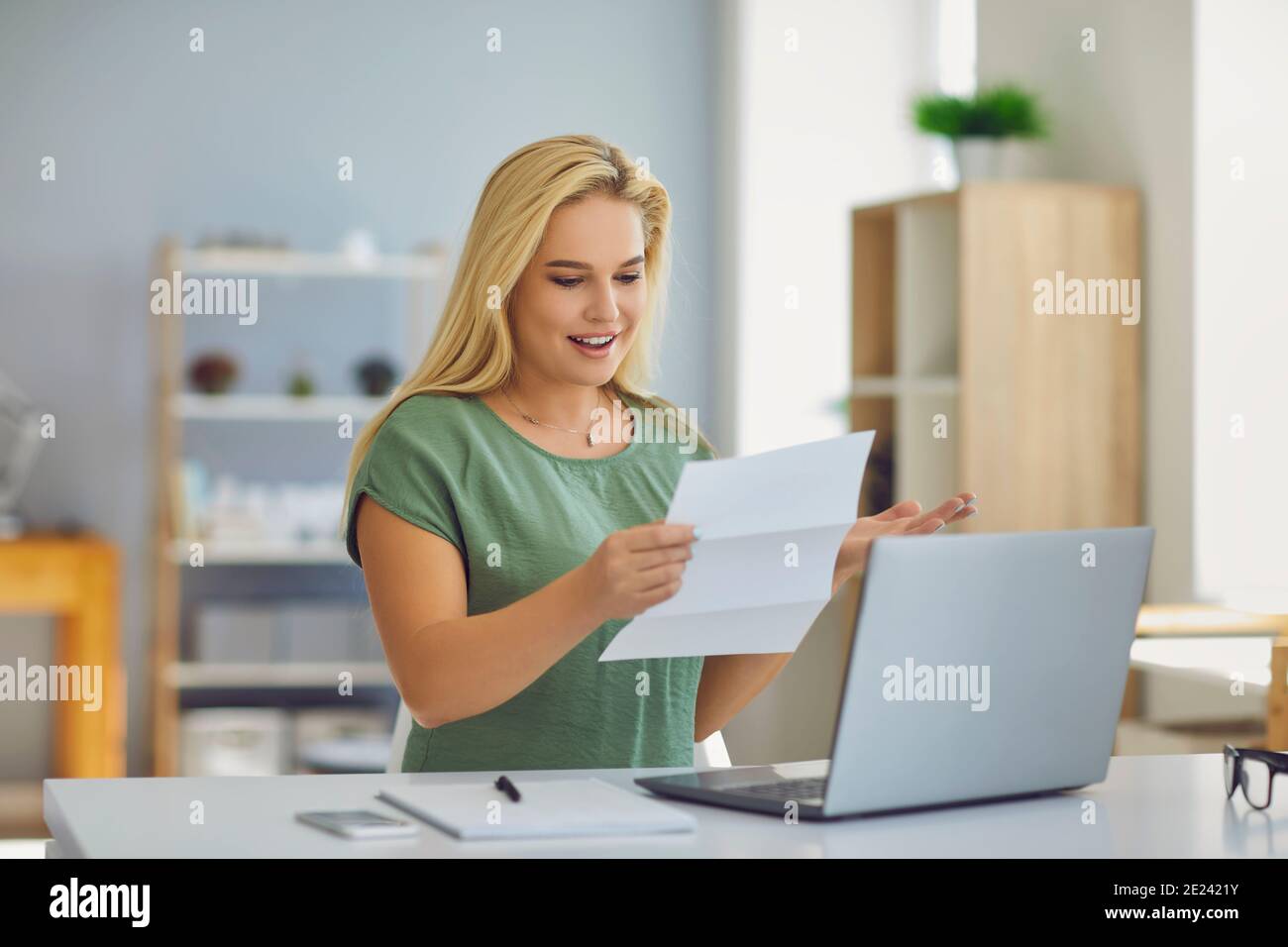 Happy young woman sitting at desk at home reading official notification ...