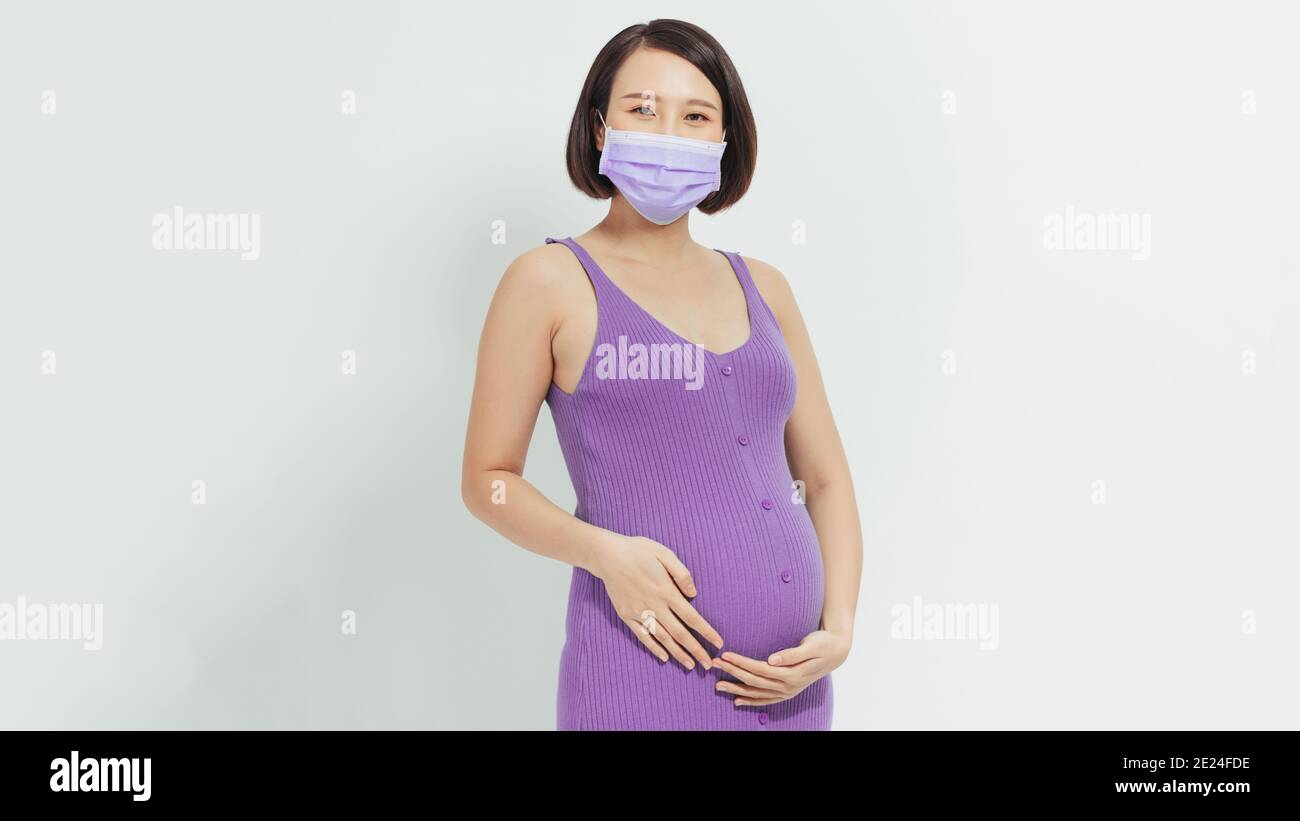 Asian pregnant woman wear face mask holding her belly, look at camera ...