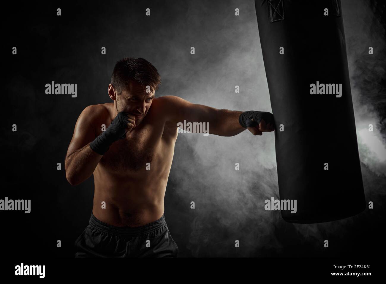 Aggressive boxer in black boxing wraps punching in boxing bag on dark ...