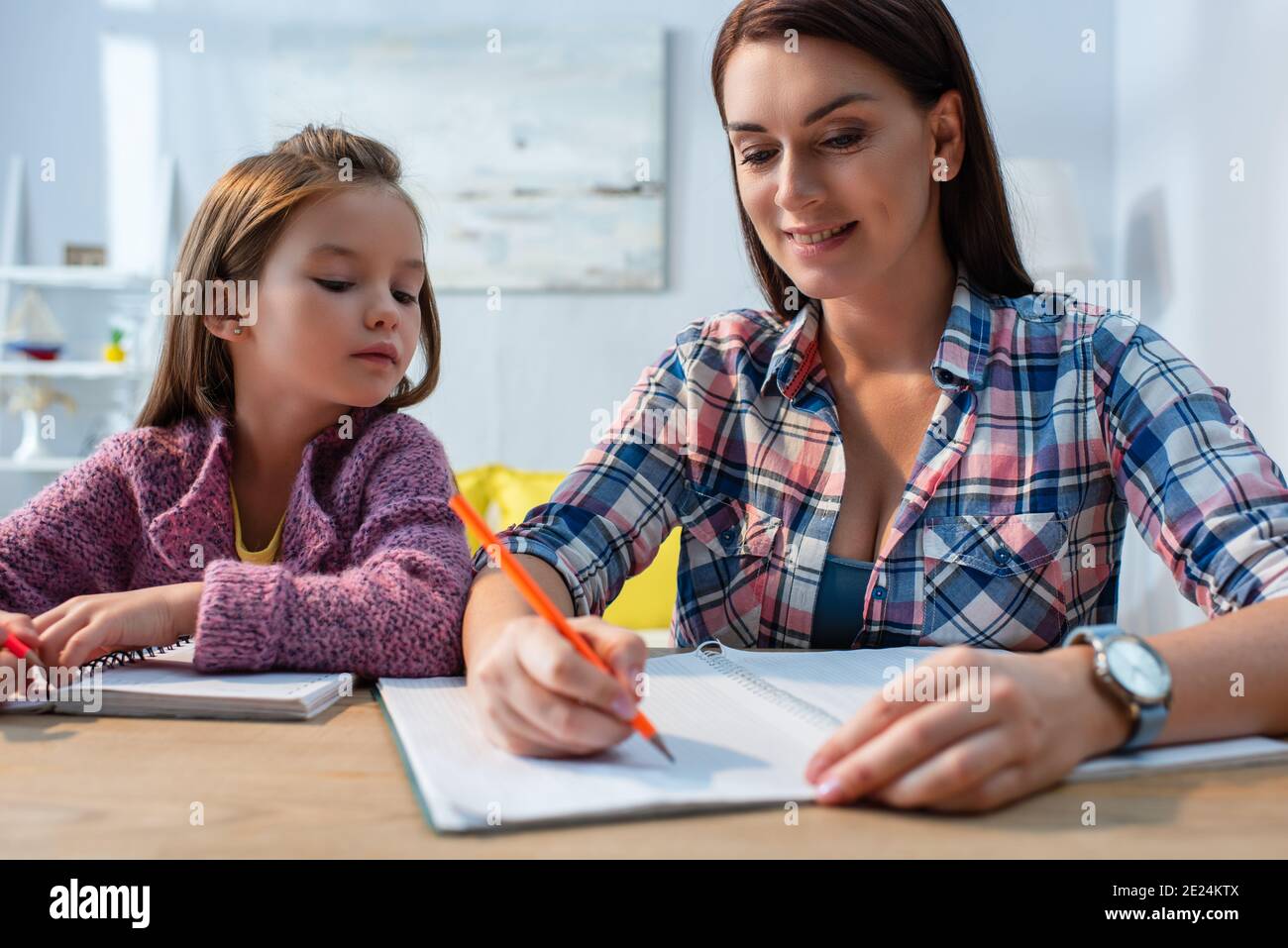 Smiling mother writing in copy book near daughter at desk on blurred ...