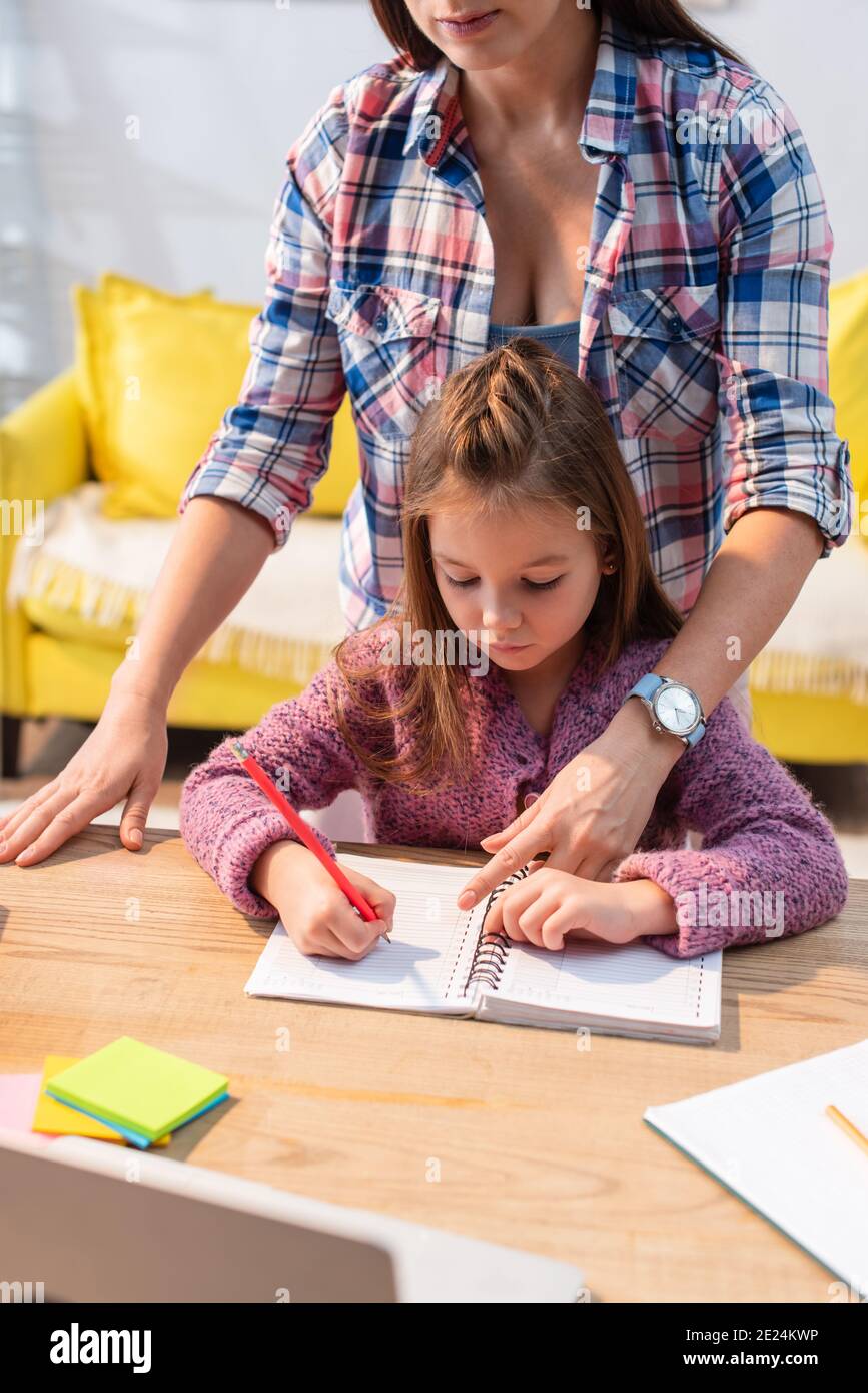 Mother pointing with finger at notebook while standing behind daughter ...