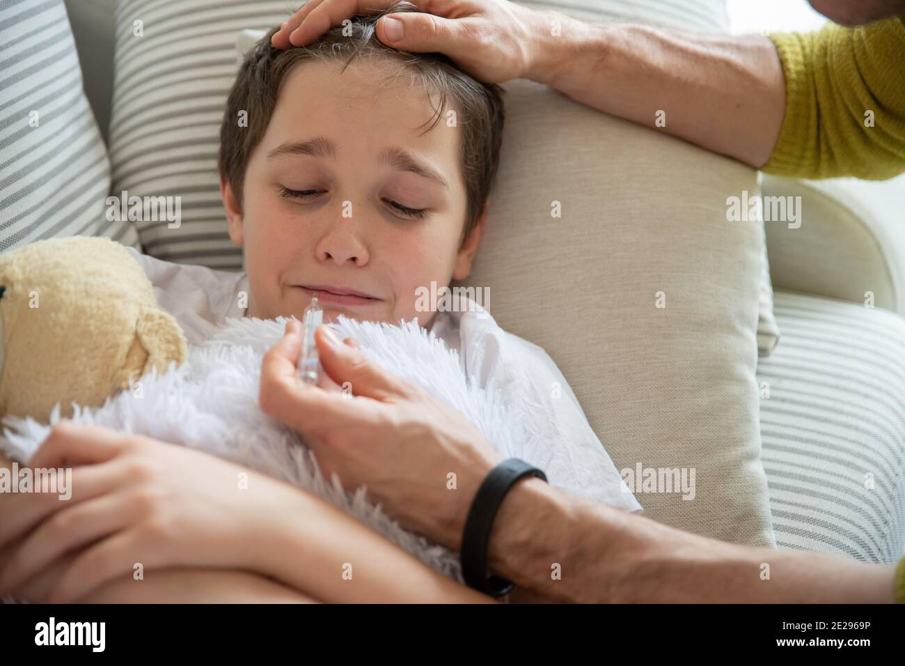 A sad crying child is lying sick on the couch. Father measures the ...