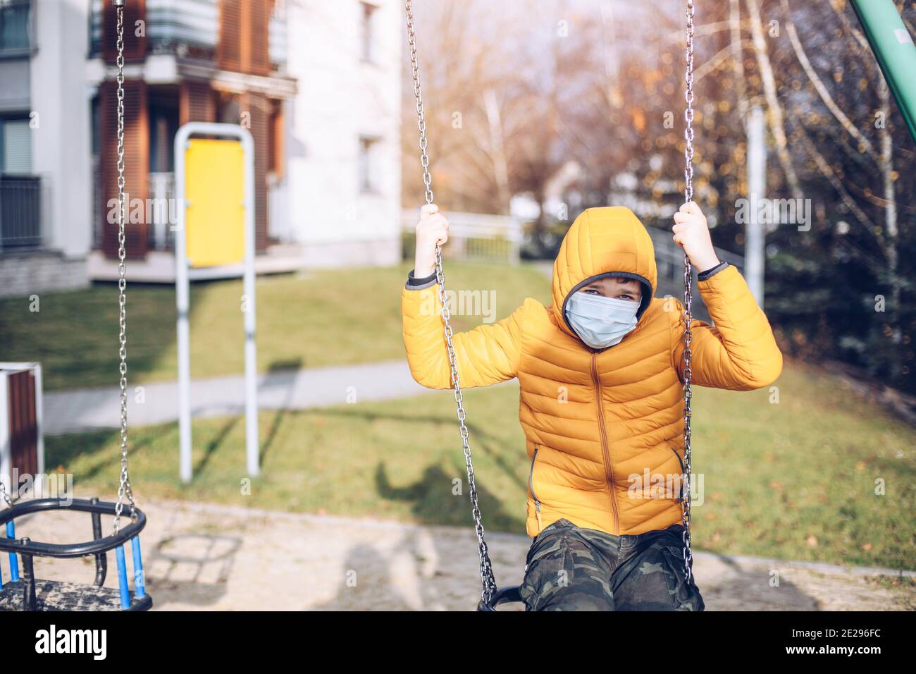 A boy in a yellow jacket and a protective mask is swinging on a swing ...