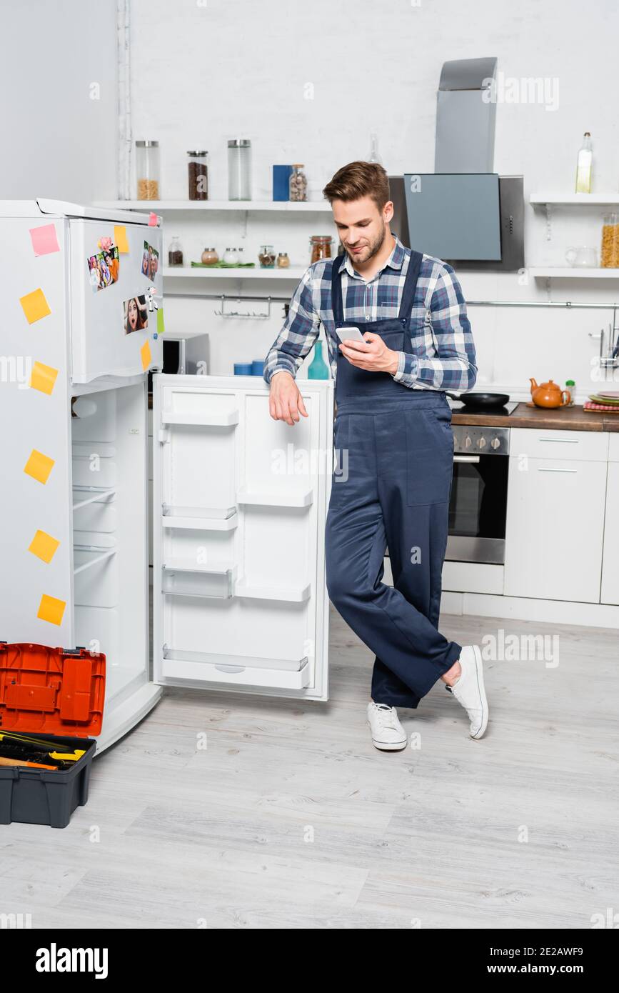 full length of smiling handyman texting on smartphone while leaning on ...