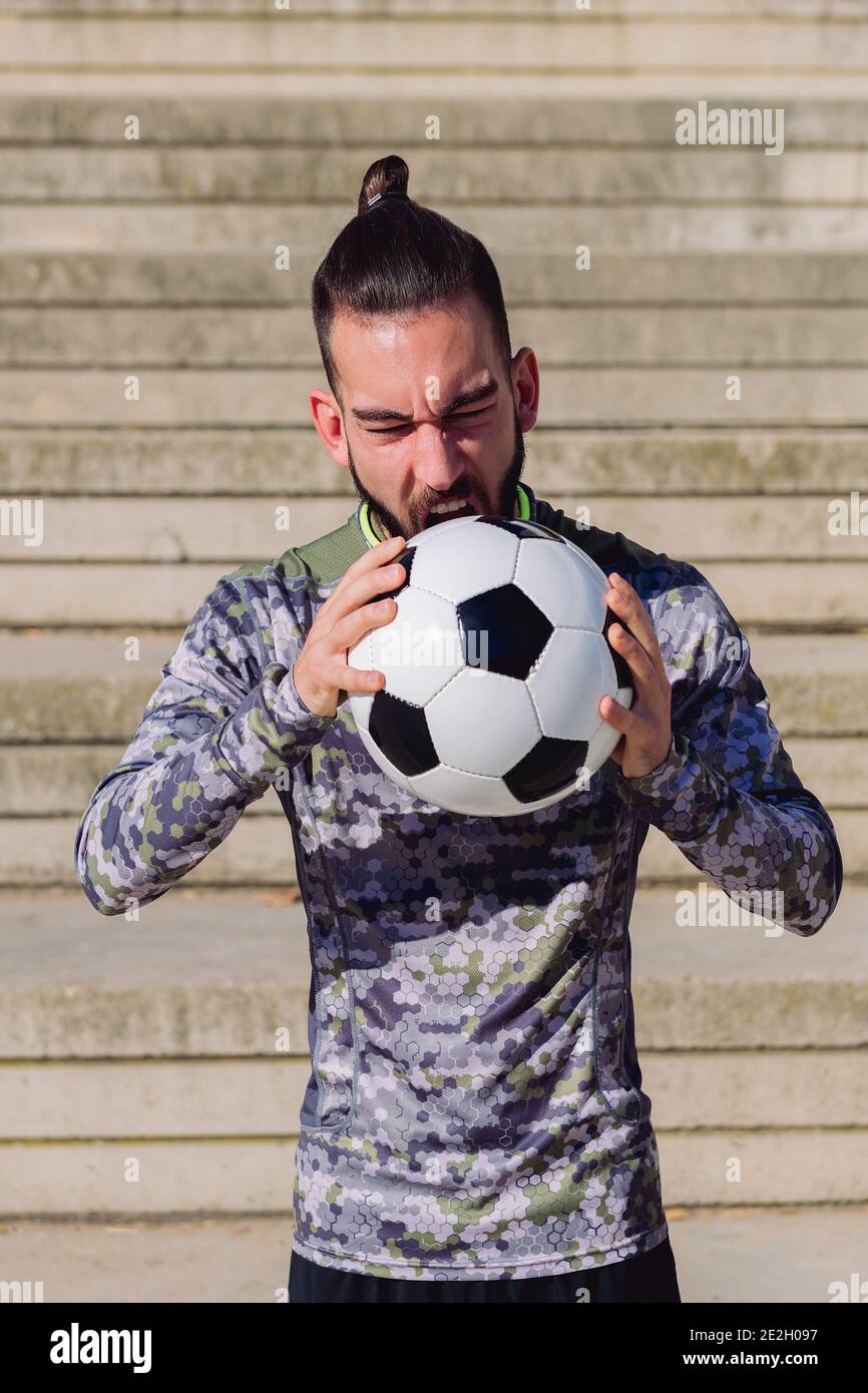 handsome sportsman in a concrete soccer court biting the football ball ...