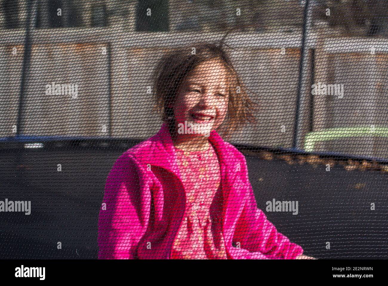 A smiling little girl with static hair sits on trampoline with net ...