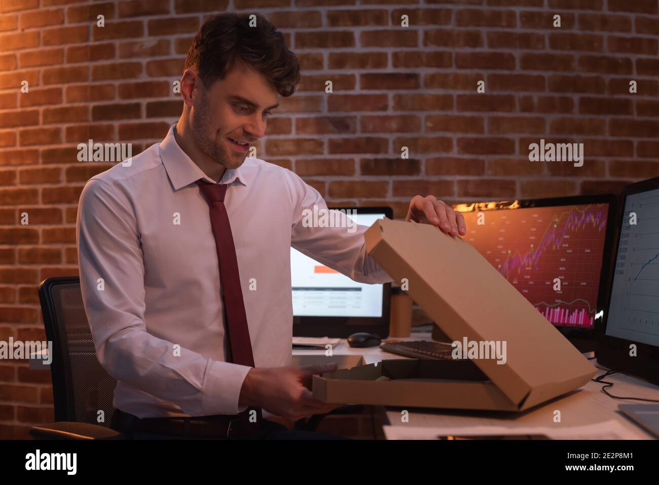 Smiling businessman opening pizza box near computers on blurred ...