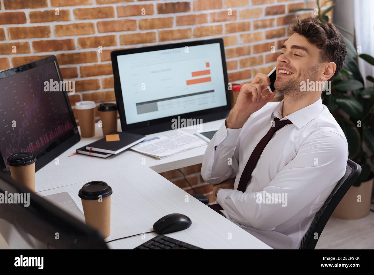 Smiling businessman talking on smartphone near computers with charts of ...