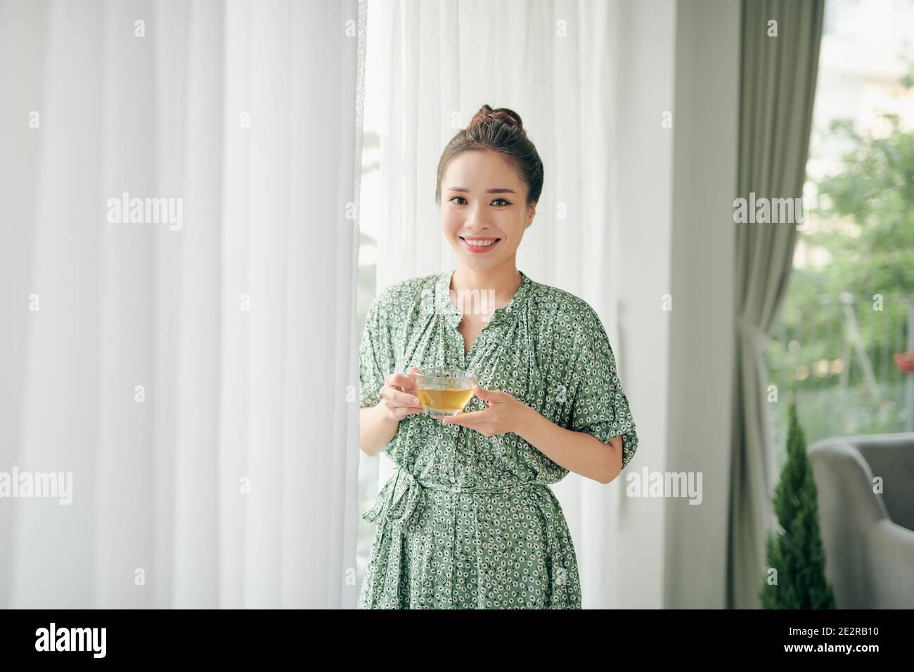 Beautiful young woman is looking out the window and holding a cup of ...