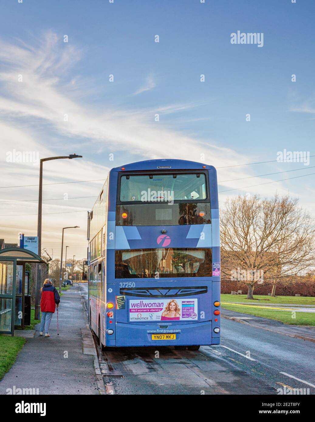 A double-decker bus stands beside a village bus stop. A woman is on the ...