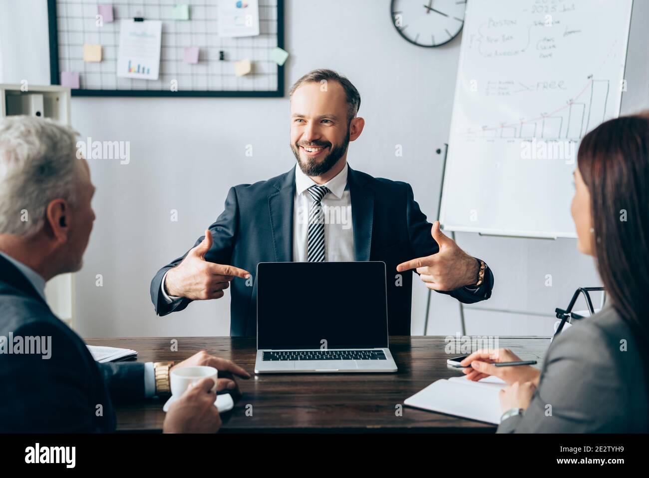 Smiling investor pointing at laptop near businesspeople with coffee and ...