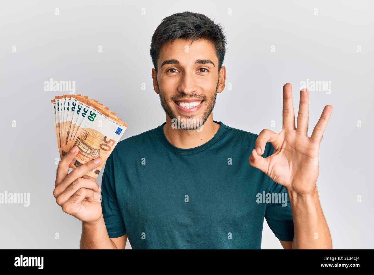 Young handsome man holding bunch of 50 euro banknotes doing ok sign ...