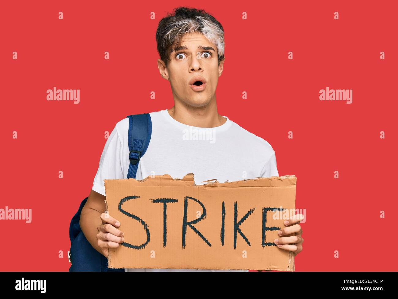Young hispanic man holding strike banner cardboard scared and amazed ...