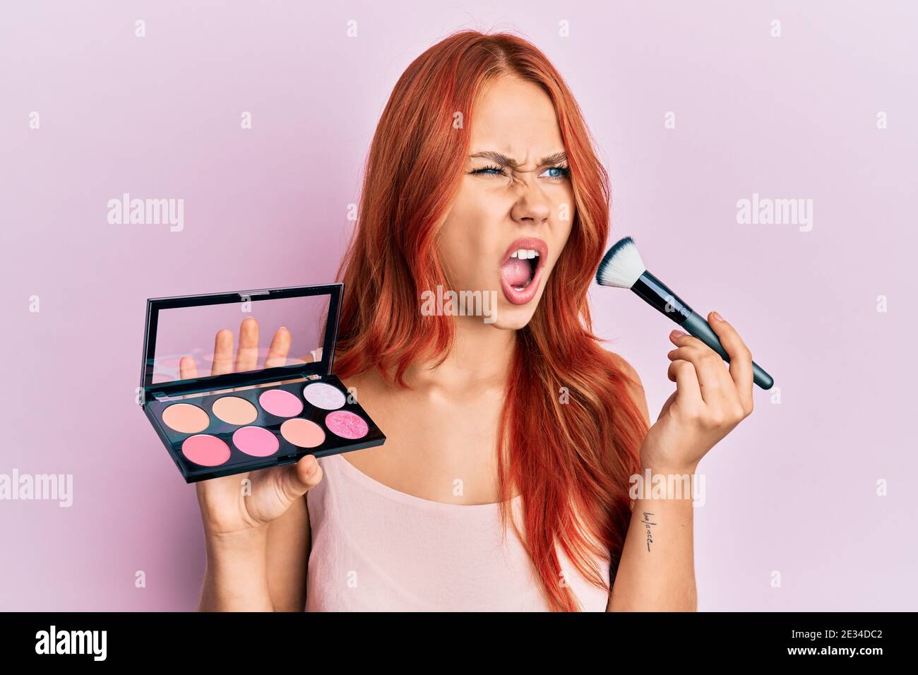 Young redhead woman holding makeup and brush angry and mad screaming ...
