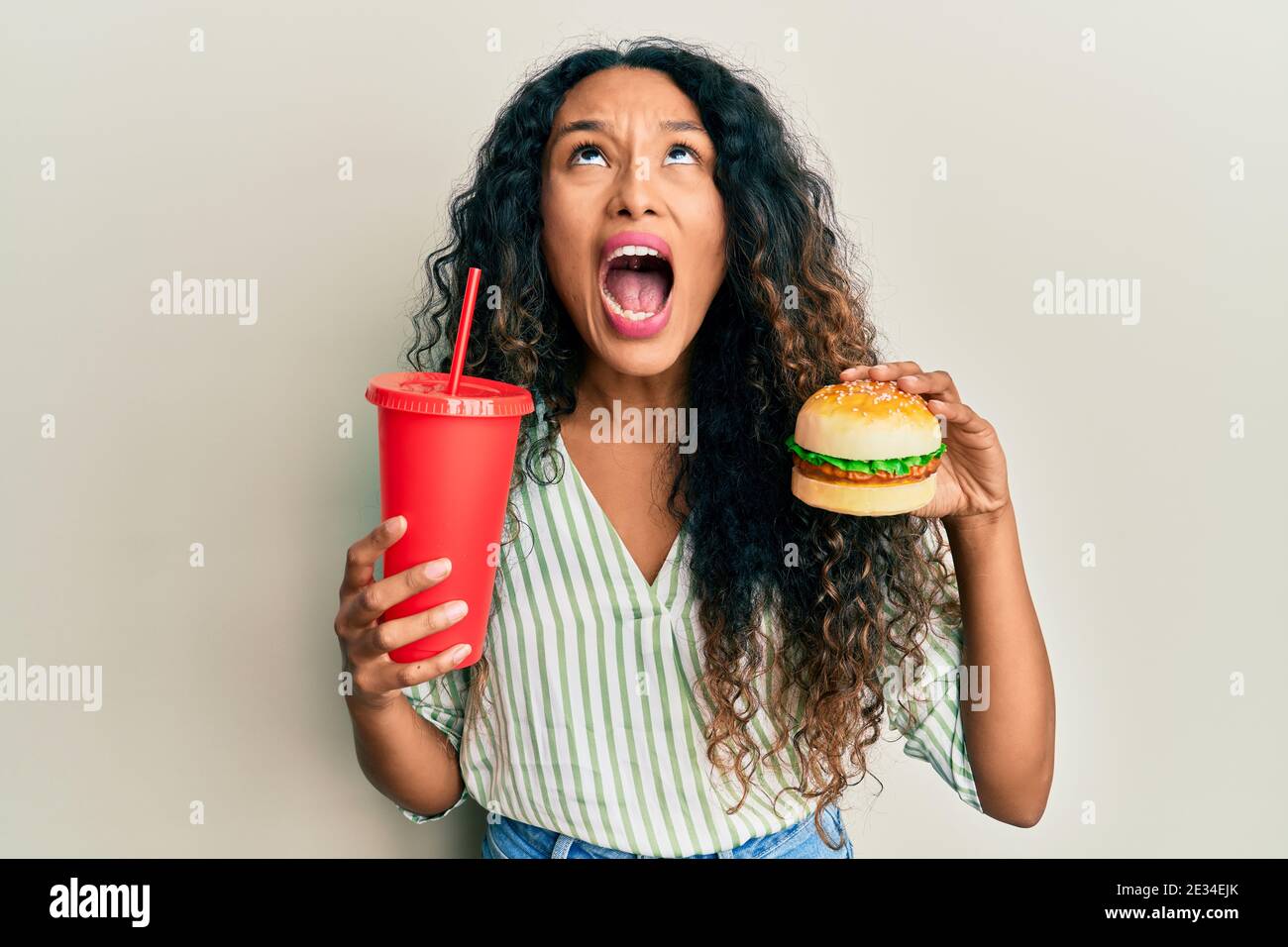 Young latin woman eating a tasty classic burger and soda angry and mad ...