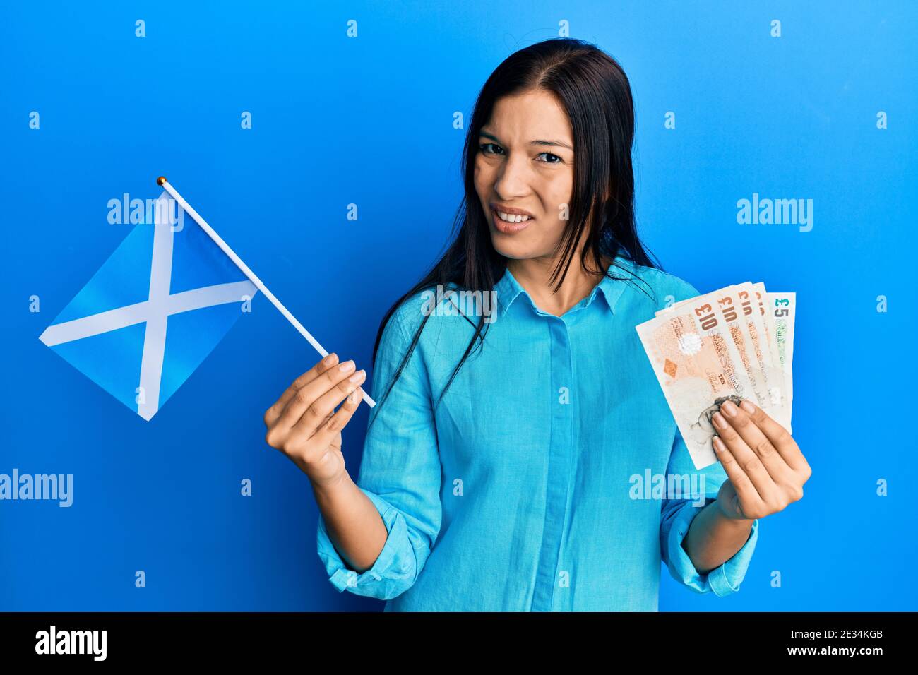 Young latin woman holding scotland flag and pounds banknotes clueless ...
