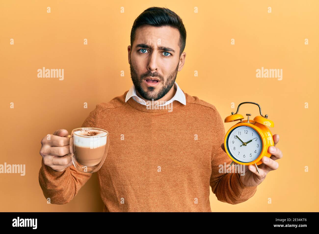 Young hispanic man holding coffee and alarm clock clueless and confused ...