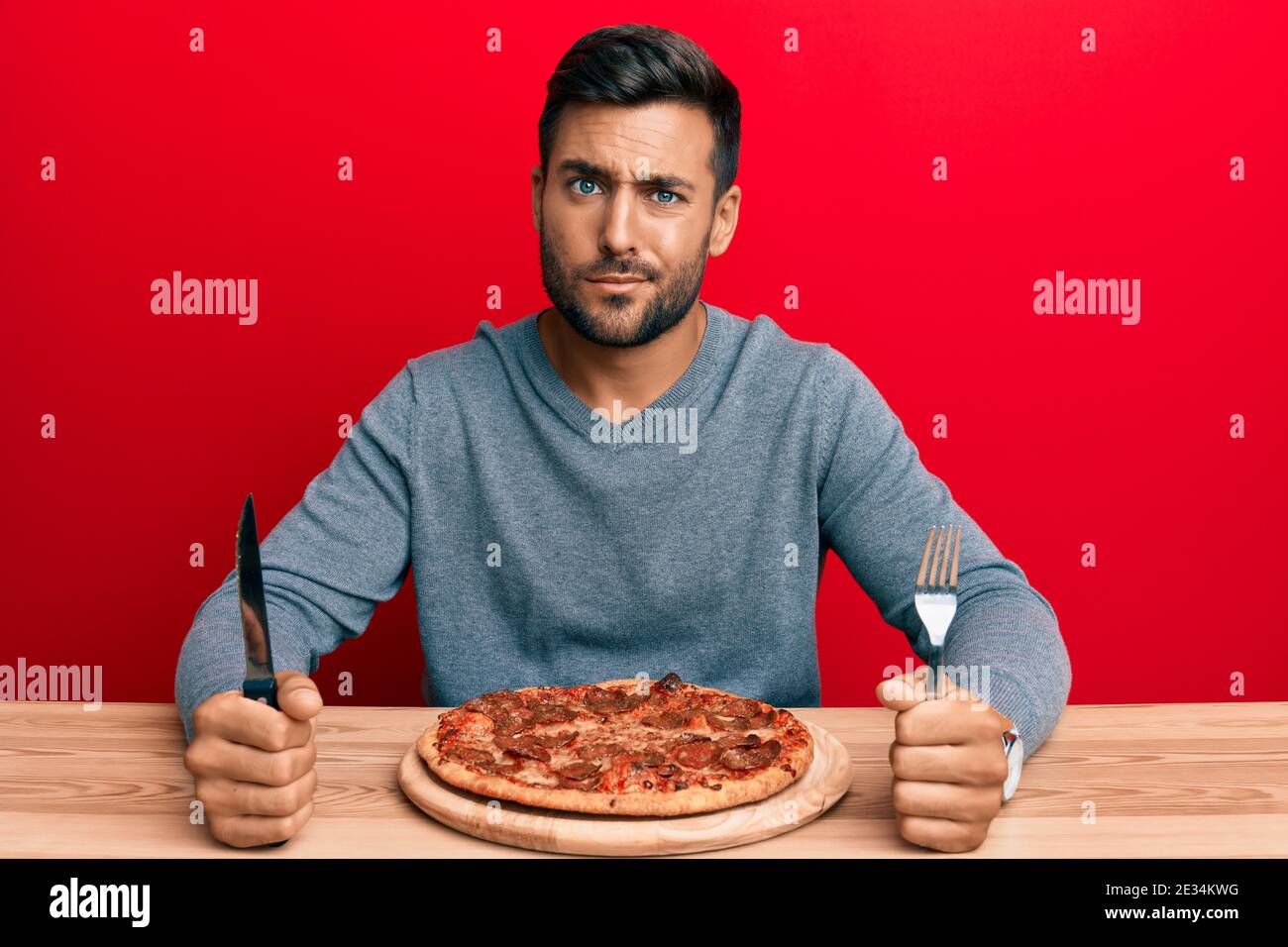 Handsome hispanic man eating tasty pepperoni pizza skeptic and nervous ...