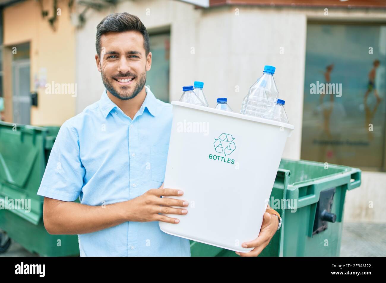 Young hispanic man smiling happy recycling. Holding full bin of plastic ...